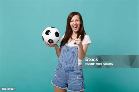 웃 고 여자 축구 팬 응원은 파란색 청록색 벽 배경에 고립 된 카메라에 축구 공을 가리키는 인덱스 손가락으로 좋아하는 팀 지원 사람들의 감정 스포츠 가족 레저 개념입니다