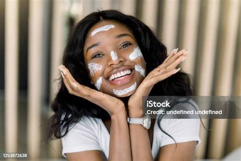 스파 스킨 케어 여성 페이셜 트리트먼트 하얀 진흙 얼굴 마스크를 바르고 카메라를 바라보며 미소짓는 즐거운 아프리카 여성의 초상화를 클로즈업하세요 아늑한 홈 인테리어 건강