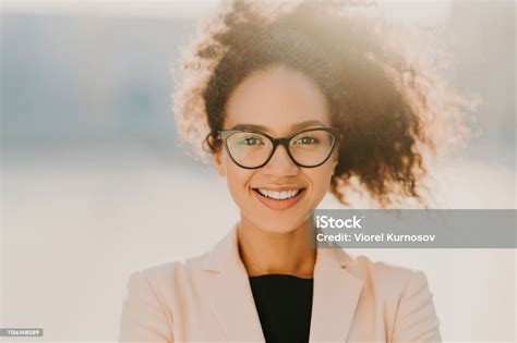 긍정적 인 표정으로 쾌활한 곱슬 머리 여자의 헤드 샷 안경을 착용 흰색 우아한 재킷 카메라를 똑바로 보고 야외에서 서 곱슬 머리는 좋은 감정을 표현한다 민족 개념 여자에