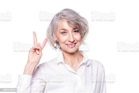 편안한 흰색 배경 위에 서있는 우아한 셔츠를 입은 선임 회색 머리 여성의 초상화를 클로즈업하여 승리의 사인을하는 카메라에서 행복한 얼굴 윙크를 하고 있습니다 여자 노인에