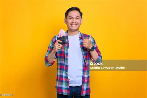 현금 돈으로 지갑을 들고 노란 배경에 고립 된 제스처를 엄지 손가락을 보여주는 격자 무늬 셔츠를 입은 쾌활한 젊은 아시아 남자 30 34세에 대한 스톡 사진 및 기타 이미지