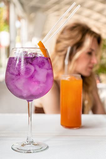 보라색으로 가득 찬 우는 유리는 매우 페리 컬러 냉각 여름 칵테일과 얼음 조각이 초점을 잃은 여성의 초상화에 맞서 테이블 위에 서 있습니다 35 39세에 대한 스톡 사진 및