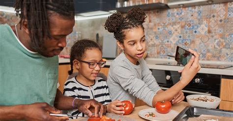 스마트폰을 들고 집에서 부엌에서 피자를 준비하는 동안 여동생과 함께 토마토를 자르는 행복한 아버지와 셀카를 만드는 웃는 다인종 소녀의 허리 위로 초상화 보기 사진