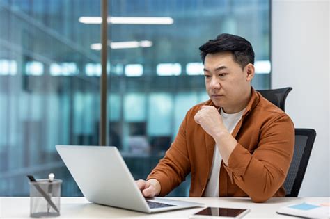 현대적인 사무실 환경에서 노트북으로 작업하는 아시아 남자 사업가 깊은 생각에 잠기고 캐주얼 한 복장을 입고 큰 창문 앞에서 집중하고 당면한 작업에 종사하고 있습니다 개발에