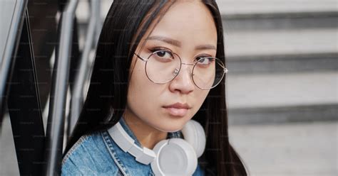 젊은 아시아 스케이터 소녀가 계단에서 롱보드에 앉아 스마트폰을 사용하고 청바지를 입고 분홍색 벨트 가방을 착용하고 둥근 안경과 흰색 헤드폰을 착용하고 있는 클로즈업