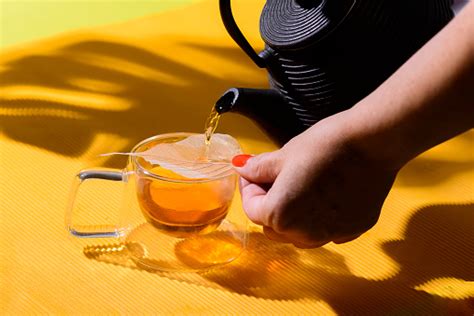 해골 잎을 차 여과기로 잡고 검은 주철 찻 주전자에서 홍차를 붓고 거친 대비 빛과 몬스테라 잎 그림자가있는 주황색 배경의 이중 벽 디자이너 유리 컵에 홍차를 붓습니다