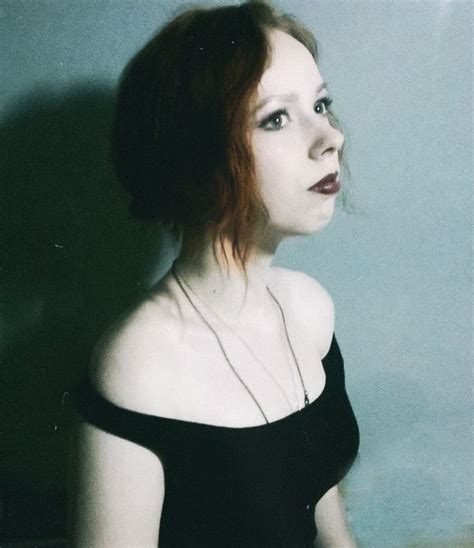 A Woman With Red Hair Wearing A Black Dress And Necklace Standing In Front Of A Blue Wall