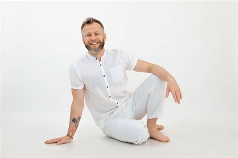 웃고 있는 잘생긴 중년 수염 난 남자가 짧은 회색 머리를 하고 흰색 배경에 바닥에 다리를 꼬고 앉아 있다 중장년에 대한 스톡