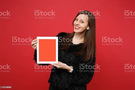 스튜디오에서 빨간 벽 배경에 고립 된 빈 빈 화면으로 태블릿 Pc 컴퓨터를 들고 검은 모피 스웨터에 웃는 젊은 여자 사람들이 진심으 감정 라이프 스타일 개념 복사 공간을