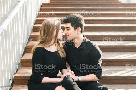아름 다운 젊은 여러 인종 커플 사랑에 학생 부부는 도시에서 나무 계단을 앉아 있다 아름 다운 터키어 갈색 머리 남자가 검은 옷에 백인 여자를 포옹 화창한 날씨에 젊은이