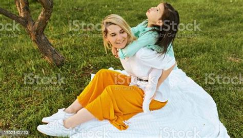 공원에서 그녀의 엄마를 포용 행복 한 아이의 측면 보기 귀여운 딸 편승 그녀의 젊은 어머니는 녹색 잔디 야외에서 흰색 담요에 앉아 어머니의 날입니다 모성 및 유년기 개념