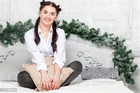 흰색 셔츠와 파란색 청바지에 젊은 아름다운 갈색 머리 여자는 크리스마스 2020 장식 방에 자신의 손에 베개와 침대에 앉아있다 2020년에 대한 스톡 사진 및 기타 이미지