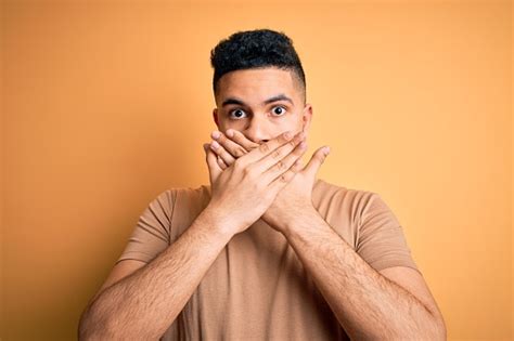 고립 된 노란색 배경 위에 서있는 캐주얼 한 티셔츠를 입은 젊은 잘생긴 남자는 실수로 손으로 입을 덮는 충격을 받았습니다 비밀 개념입니다 T 셔츠에 대한 스톡 사진 및 기타