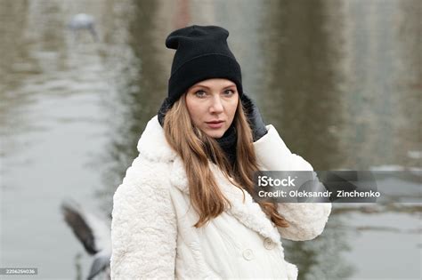 한 젊은 여성이 포근한 흰색 코트와 검은색 비니를 쓴 채 고요한 수원 옆에 서 있습니다 그녀의 긴 머리칼이 어깨 너머로 흘러내리며 사려 깊은 마음으로 먼 곳을 응시한다