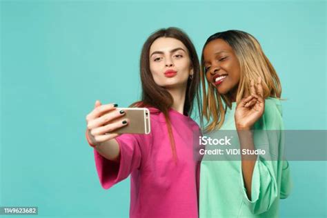 파란색 벽 배경 스튜디오 초상화에 고립 된 핸드폰에서 Selfie 샷을 하 고 분홍색 녹색 옷 두 여자 친구 유럽과 아프리카계 미국인 사람들 라이프 스타일 개념입니다 복사