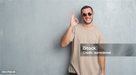 손과 손가락으로 Ok 사인을 하 고 긍정적인 미소 선글라스 입고 회색 그런 지 벽에 젊은 백인 남자 성공적인 식입니다 그런지 이미지 기법에 대한 스톡 사진 및 기타 이미지