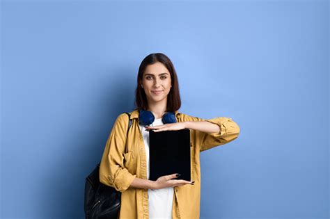 긍정적 인 자신감학생 소녀는 손으로 태블릿을 들고 노란색 셔츠 흰색 티셔츠 검은 색 가방과 헤드폰을 목에 착용 제품 이나 서비스의 광고와 화면을 보여주는 태블릿에 대한 스톡