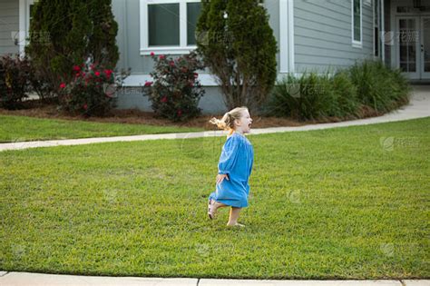 小女孩扎着辫子，穿着蓝色的裙子，在一所新建筑房子的草坪上奔跑