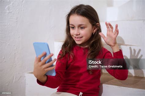 귀여운 쾌활한 어린 소녀 소녀가 스마트 폰으로 화상 통화를하는 친구 또는 친척을 맞이하고 흰 벽을 배경으로 아늑한 집의 계단에