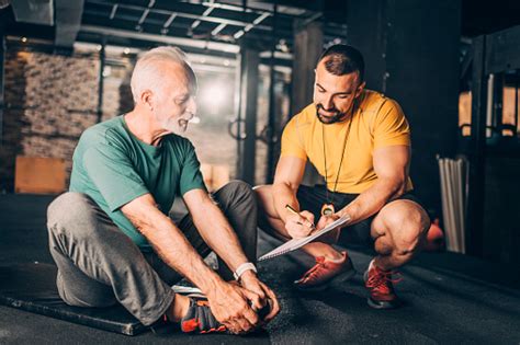 크로스 핏 체육관 바닥에 웅 크 리고 활동적인 노인 남성 운동 계획을 보여주는 개인 피트 니스 어시스턴트 2명에 대한 스톡 사진 및 기타 이미지 Istock