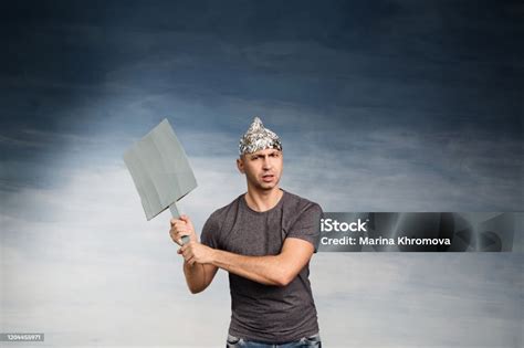 호일 모자를 쓴 괴짜 남자가 무서워서 손으로 손을 잡는다 넓은 눈과 입을 열고 파란색 배경에 스트라이크 포스터를 들고 있습니다 Popsicle Stick에 대한 스톡 사진