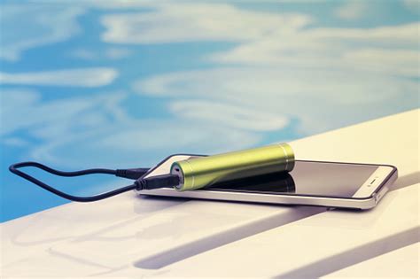파란색 물 배경의 Usb 케이블을 통해 녹색 휴대용 배터리 충전기에 연결된 스마트 폰 나머지 기간 동안 수영장 근처 의 라운저에 전화의 비상 충전 무선 기술에 대한 스톡