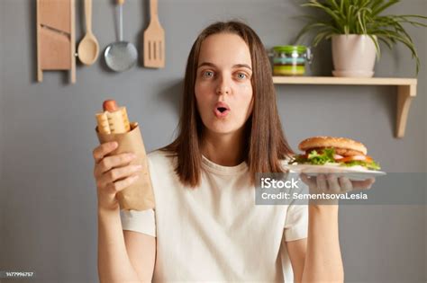 맛있는 햄버거와 핫도그를 먹고 다이어트를 깨고 패스트 푸드를 좋아하고 흰색 티셔츠를 입고 카메라를보고 와우라고 말하면서 놀란 여자는 놀란 표정을 짓습니다 30 34세에 대한