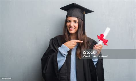 손과 손가락으로 가리키는 매우 행복도 들고 대학원 제복을 입고 그런 지 회색 벽에 젊은 갈색 머리 여자 가리키기에 대한 스톡