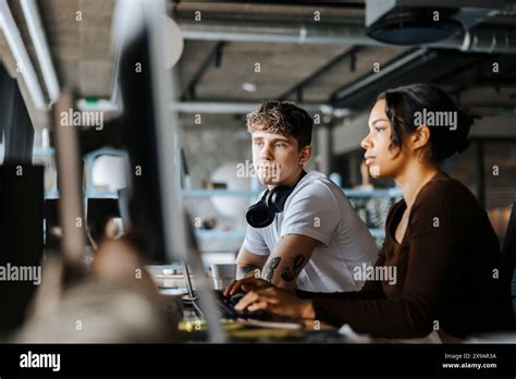 Young Male Computer Programmer Sitting Next To Female Colleague Working On Computer At Desk In