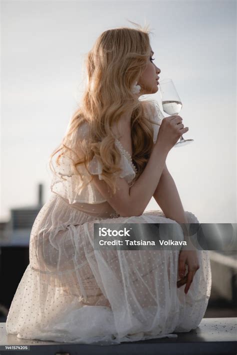 낭만적인 흰색 드레스를 입은 아름다운 여자가 옥상 테라스에서 샴페인 한 잔을 마시며 옆으로 쪼그리고 앉아 시선을 돌린다 세련된 복장과 완벽한 헤어 스타일의 우아한 숙녀 파리