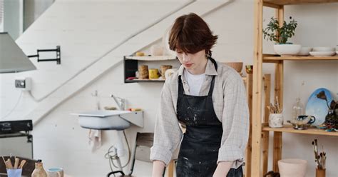 검은 앞치마를 입은 젊은 공예가 또는 토기 가게 주인이 테이블 옆에 서서 밀대로 점토 조각을 평평하게 한다 사진
