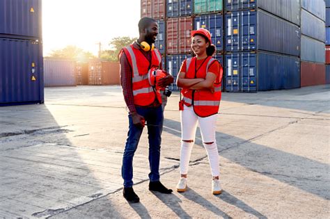 두 명의 행복한 미소 산업 아프리카 엔지니어 남자와 여자는 안전 조끼와 헬멧을 착용하고 황혼 일몰 시간에 물류 선적화물 컨테이너