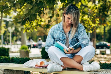 아름다운 여성의 수평 야외 이미지는 거리에서 화창한 날에 벤치에 앉아있는 동안 책을 읽습니다 청바지 재킷을 입은 예쁜 여성이 밖에서 휴식을 취하고 책에서 배운다 30 39