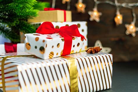 빨간 리본 리본으로 묶인 흰색과 금색 선물 상자무리가 크리스마스 트리 아래에 빛나는 화환이 있는 어두운 나무 벽에 놓여 있습니다 사이드 뷰 클로즈업 새해 구성 크리스마스에