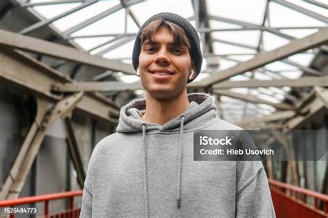 회색 후드티와 흰색 무선 이어버드 헤드폰이 달린 모자를 쓰고 웃는 힙스터 남자의 도시 초상화 남자에 대한 스톡 사진 및 기타 이미지 Istock