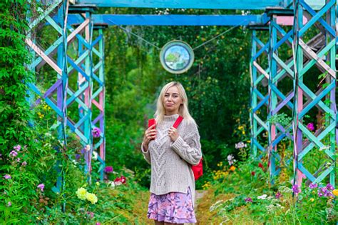 빨간 배낭을 등에 메고 있는 니트 스웨터를 입은 어린 금발 소녀가 손에 든 전화가 공원이나 숲의 나무 색 아치의 길을 따라 걷고 있습니다 야생 여행 관광에 대한 스톡 사진