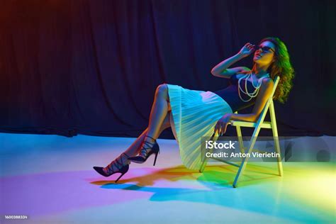 하얀 의자에 앉아 네온 불빛의 짙은 보라색 배경 위로 머리를 뒤로 젖히는 세련된 의상을 입은 젊은 여성의 가로 이미지 고급 의상에 대한 스톡 사진 및 기타 이미지 Istock