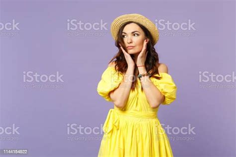 노란 드레스에 멋진 젊은 여자 여름 모자는 스튜디오에서 파스텔 바이올렛 배경에 고립 된 옆을 보고 턱에 손을 소품을 넣어 사람들은 진심으로 감정 라이프 스타일 개념입니다