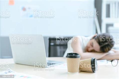 피곤한 비즈니스 아시아 여성이 배경에서 자고 있는 테이블 위에 두 개의 커피 종이컵이 있습니다 초과 근무 개념입니다 종이컵에 초점을 선택합니다 두통에 대한 스톡 사진 및