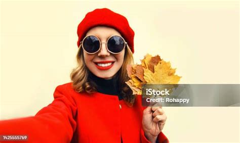 배경에 빨간 프랑스 베레를 착용 한 노란색 단풍 나무 잎이있는 스마트 폰으로 셀카를 찍기 위해 손을 뻗고있는 아름다운 웃는 여성의 가을 초상화 25 29세에 대한 스톡 사진