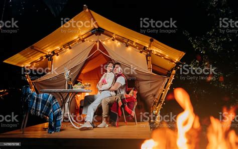 여름 저녁에 글램핑에서 휴식을 취하고 아늑한 모닥불 근처에서 와인을 마시는 행복한 사랑스러운 커플 야외 레크리에이션 및 레크리에이션을 위한 고급 캠핑 텐트 라이프 스타일