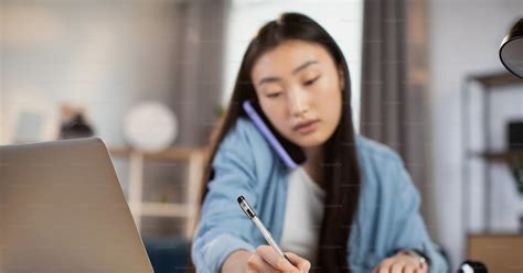 매력적인 아시아 중국 여성은 집에서 직장에서 현대적인 노트북으로 앉아 모바일로 이야기하고 메모를 작성합니다 가제트와 통신의 개념 펜으로 손에 집중 사진