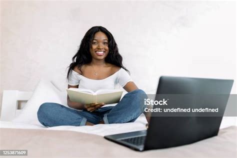 흰색 티셔츠와 청바지를 입은 젊은 아프리카 계 미국인 소녀는 침실에서 노트북 Pc와 메모장이있는 침대에 앉아 집에서 공부하거나