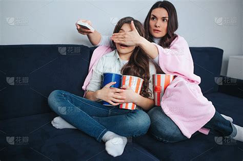 美丽的黑发白人母亲和女儿一起坐在房间里。妈妈遮住了女孩的眼睛。她拿着遥控器，厌恶地期待着。女儿拿着爆