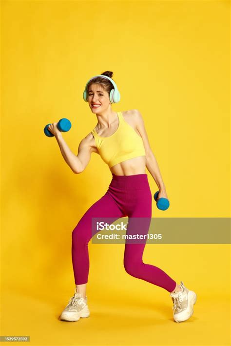 운동복을 입고 헤드폰을 끼고 노란 스튜디오 배경에 무거운 덤벨을 들고 훈련하는 젊은 날씬한 여성의 전체 길이 이미지 18 19세에 대한 스톡 사진 및 기타 이미지 Istock