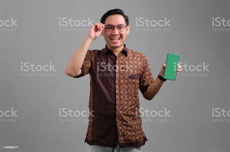 빈 화면 스마트폰을 보여주는 바틱 셔츠를 입고 회색 배경에 격리된 안경을 만지는 쾌활한 젊은 아시아 남자 30 34세에 대한 스톡 사진 및 기타 이미지 Istock