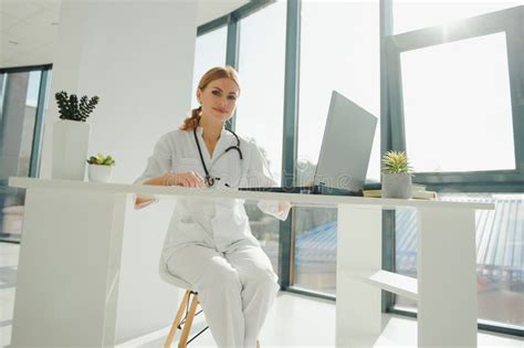 Bella Joven Y Sonriente Doctora Sentada En El Escritorio Y Escribiendo Foto De Archivo Imagen