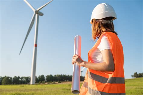 하드 햇과 보호 조끼를 입은 엔지니어는 풍차의 배경에 대해 현장에서 풍력 터빈을 건설하기위한 계획 프로젝트를 보유하고 있습니다 전기의 생성 재생 가능한 녹색 에너지의 대체