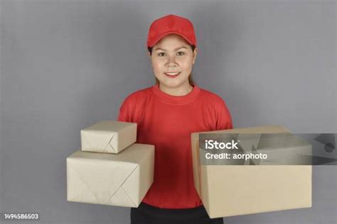 회색 배경에 소포 상자를 들고 서 있는 빨간 유니폼을 입은 행복한 배달 아시아 여성 30 34세에 대한 스톡 사진 및 기타 이미지 30 34세 개념 동남아시아 민족