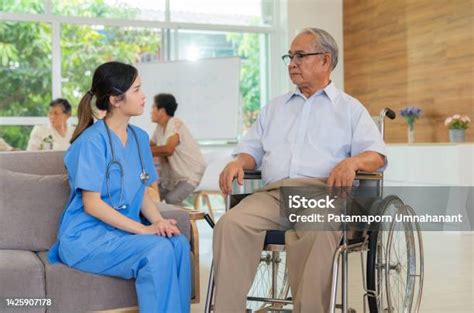 젊은 아시아 간병인 의사는 양로원의 거실에서 휠체어를 타고 노인 옆에 앉아 있습니다 정신 관리와 건강 노인을 돌보는 간호사와 미소로 물리 치료 노인에 대한 스톡 사진 및